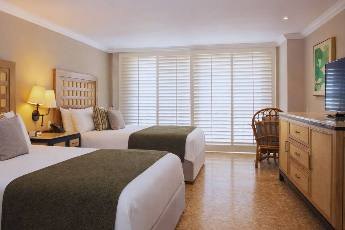 Quarto de hotel com duas camas, uma escrivaninha, uma cadeira, televisão e janela com persianas.