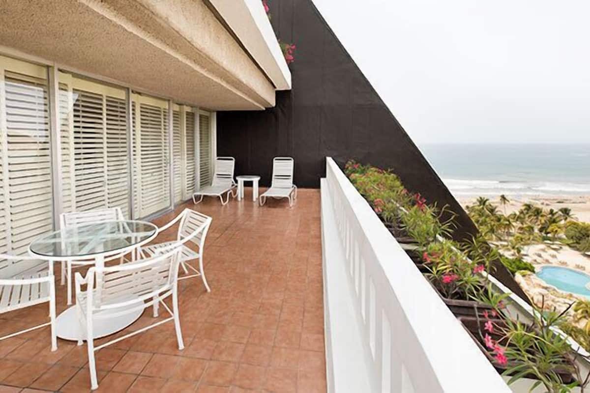 Uma varanda com mesa de vidro e cadeiras brancas, com vista para uma praia e uma piscina.