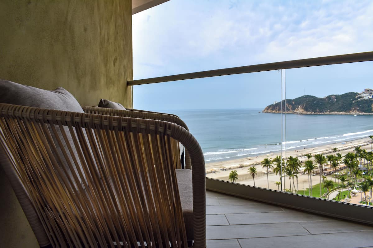 Uma cadeira de vime com almofadas, vista para a praia e uma colina distante com edifícios.
