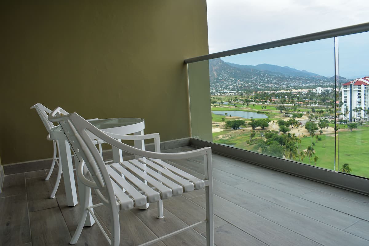 Uma varanda com cadeiras brancas e uma mesa pequena, com vista para um horizonte urbano com edifícios e um lago.