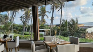 Uma área de refeições com mesa posta para quatro, com vista para uma praia com palmeiras e céu claro.