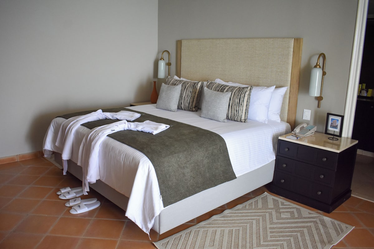 Uma cama bem arrumada com almofadas, uma mesinha de cabeceira com telefone e porta-retrato, e chinelos no chão.