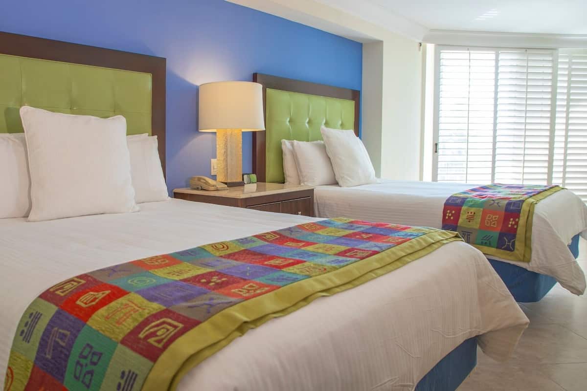 Quarto de hotel com duas camas, cada uma com roupa de cama branca e um edredom colorido. Há uma mesinha de cabeceira com uma luminária e um telefone entre as camas. O quarto tem uma parede azul e uma janela com persianas.