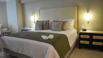 Quarto de hotel com uma cama grande, uma mesa de cabeceira com abajur e uma mesinha de cabeceira com gavetas.