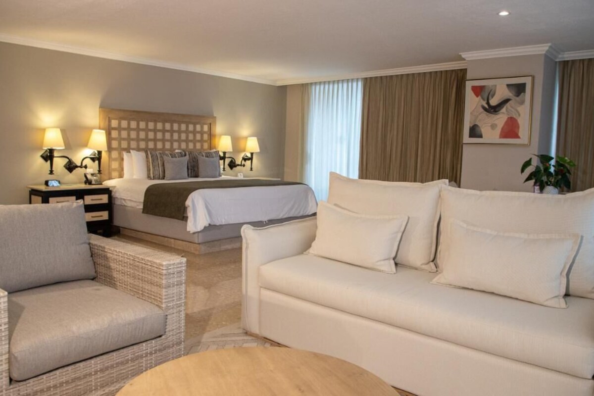 Quarto de hotel com cama, duas mesas de cabeceira com luminárias, um sofá, uma poltrona e uma janela grande com cortinas.
