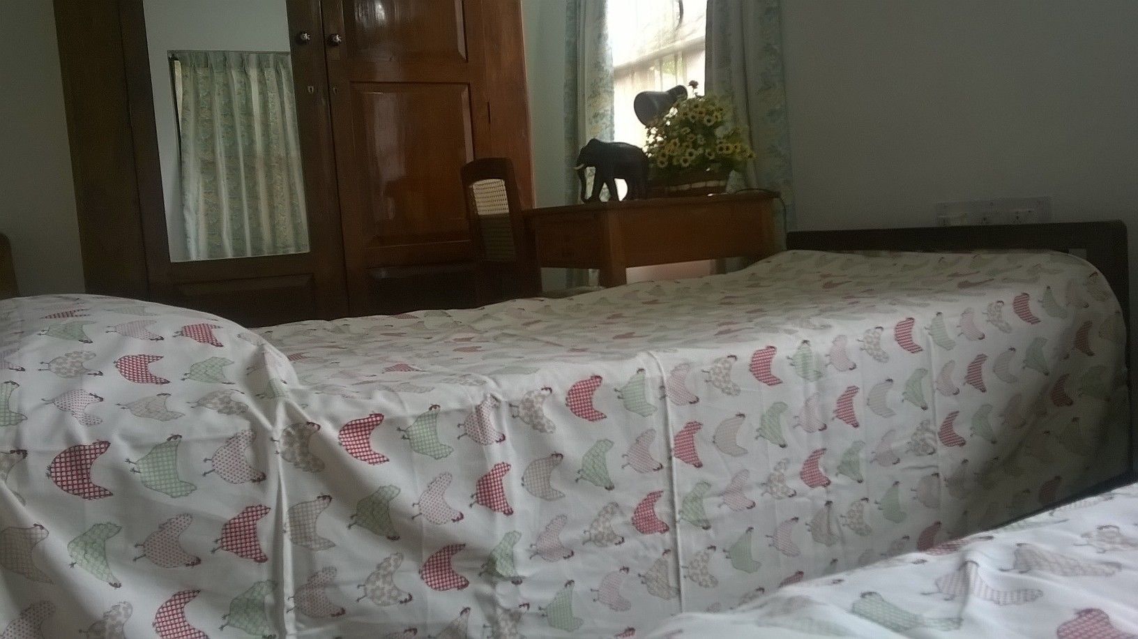 Quarto com cama coberta por um cobreleito estampado, mesa de cabeceira de madeira com luminária e flores, e um guarda-roupa.