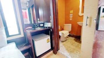 Quarto de hotel compacto com uma cama, uma pequena mesa com telefone e computador, um sanitário e uma pia.
