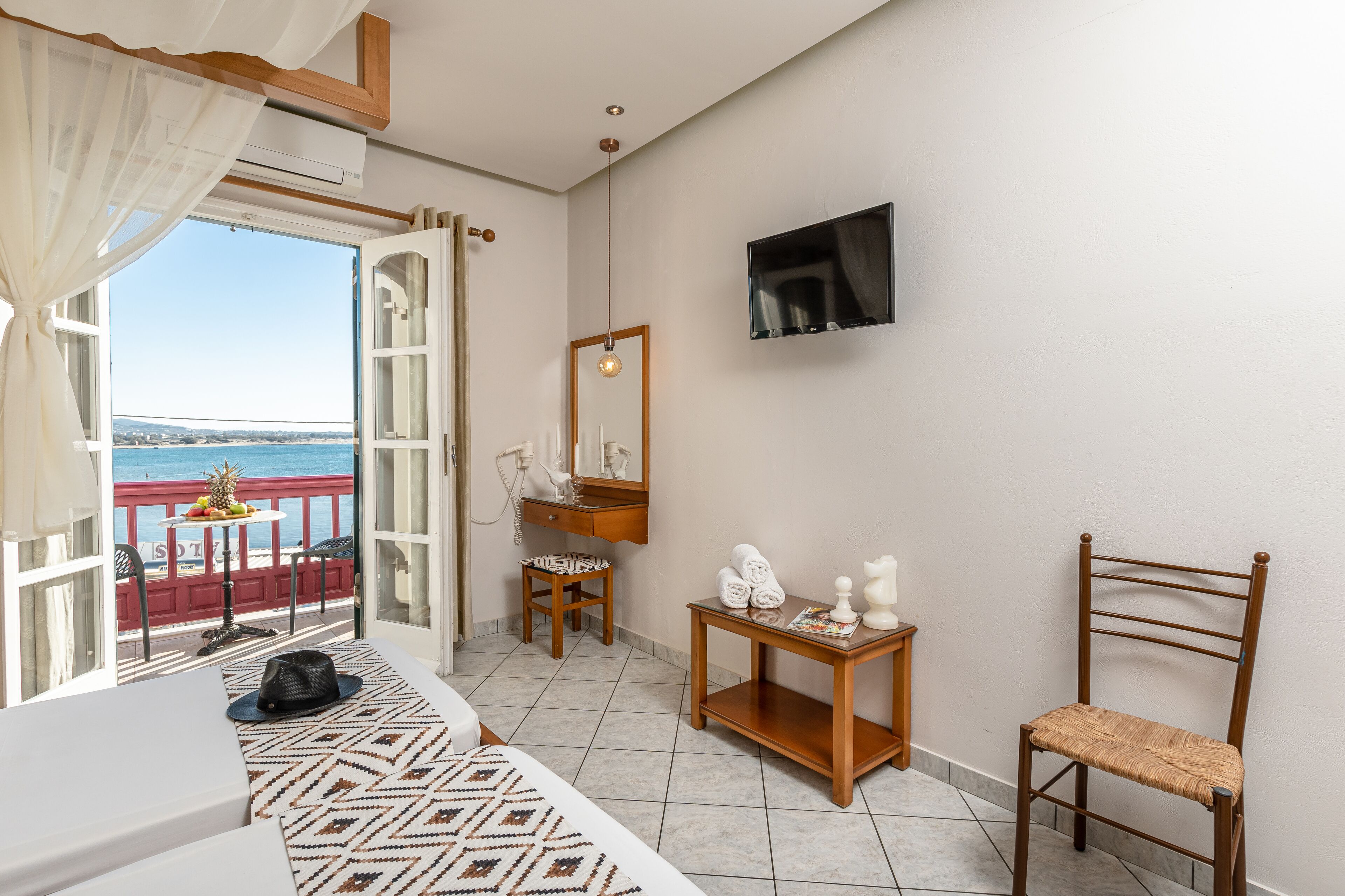 Quarto de hotel com varanda, uma cama, uma pequena mesa com um chapéu, uma cadeira de madeira, um espelho e uma TV na parede.