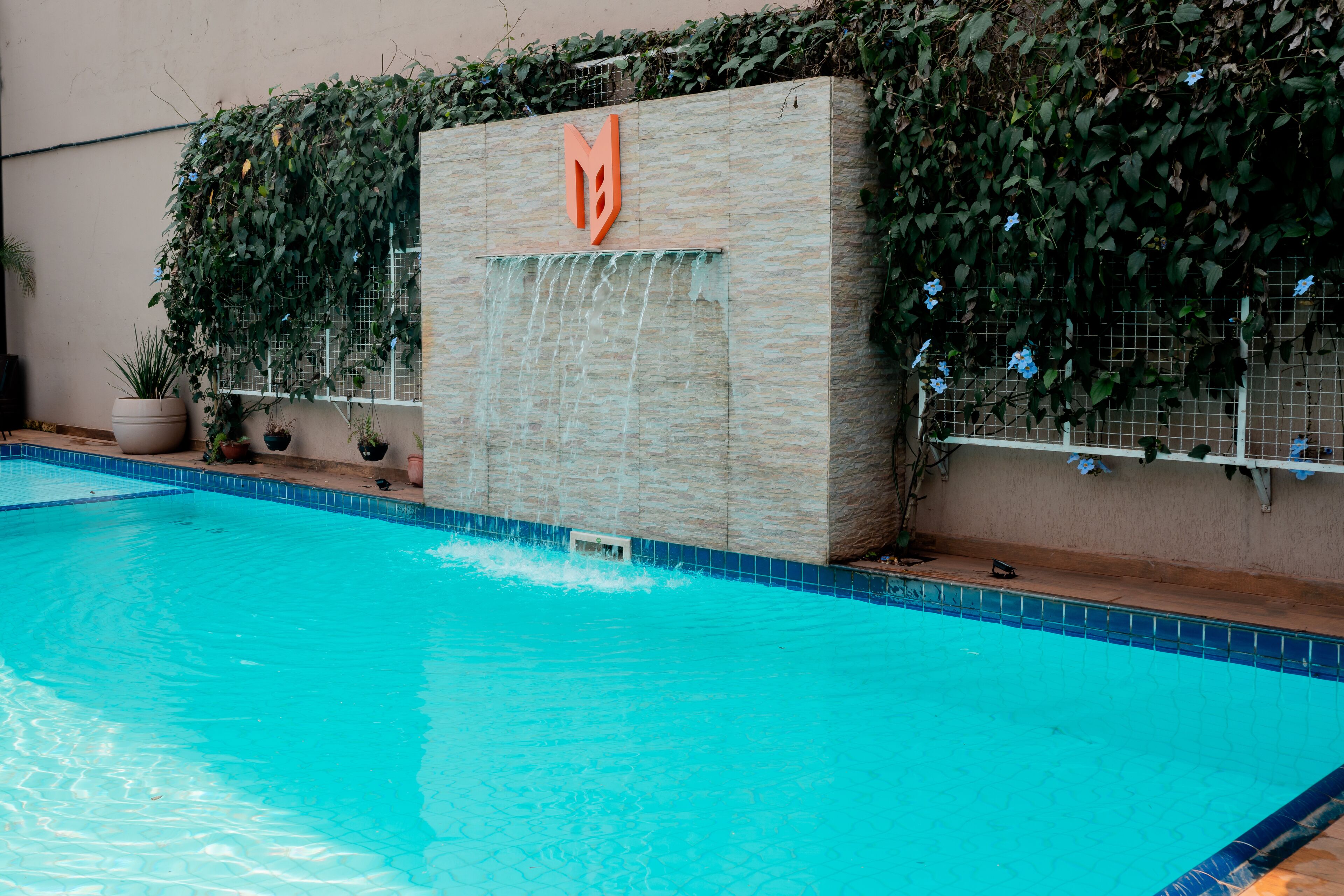 Piscina com muro de pedra e um logotipo, rodeada por plantas em vasos e uma cerca de metal.