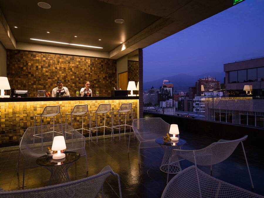 Um bar no terraço com vista para a cidade, com balcão, área de estar com mesas e cadeiras, e iluminação ambiente.