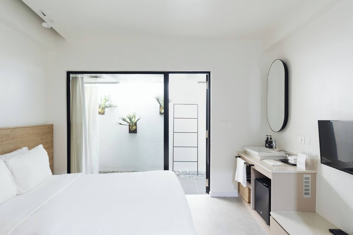 Quarto de hotel moderno com uma cama grande, banheiro com pia e espelho, e vista para um jardim por uma porta aberta.