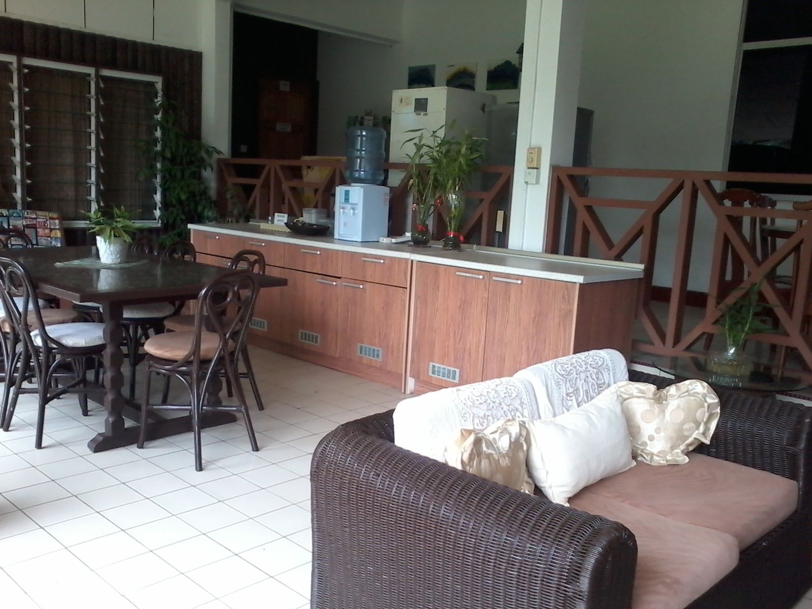 Uma área de refeições com uma mesa e cadeiras de madeira, uma cozinha com dispensador de água e armários, e um sofá de vime com almofadas.