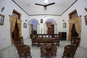 Quarto com sofá de madeira, duas cadeiras, uma mesa redonda com um arranjo floral, um ventilador de teto e quadros nas paredes.