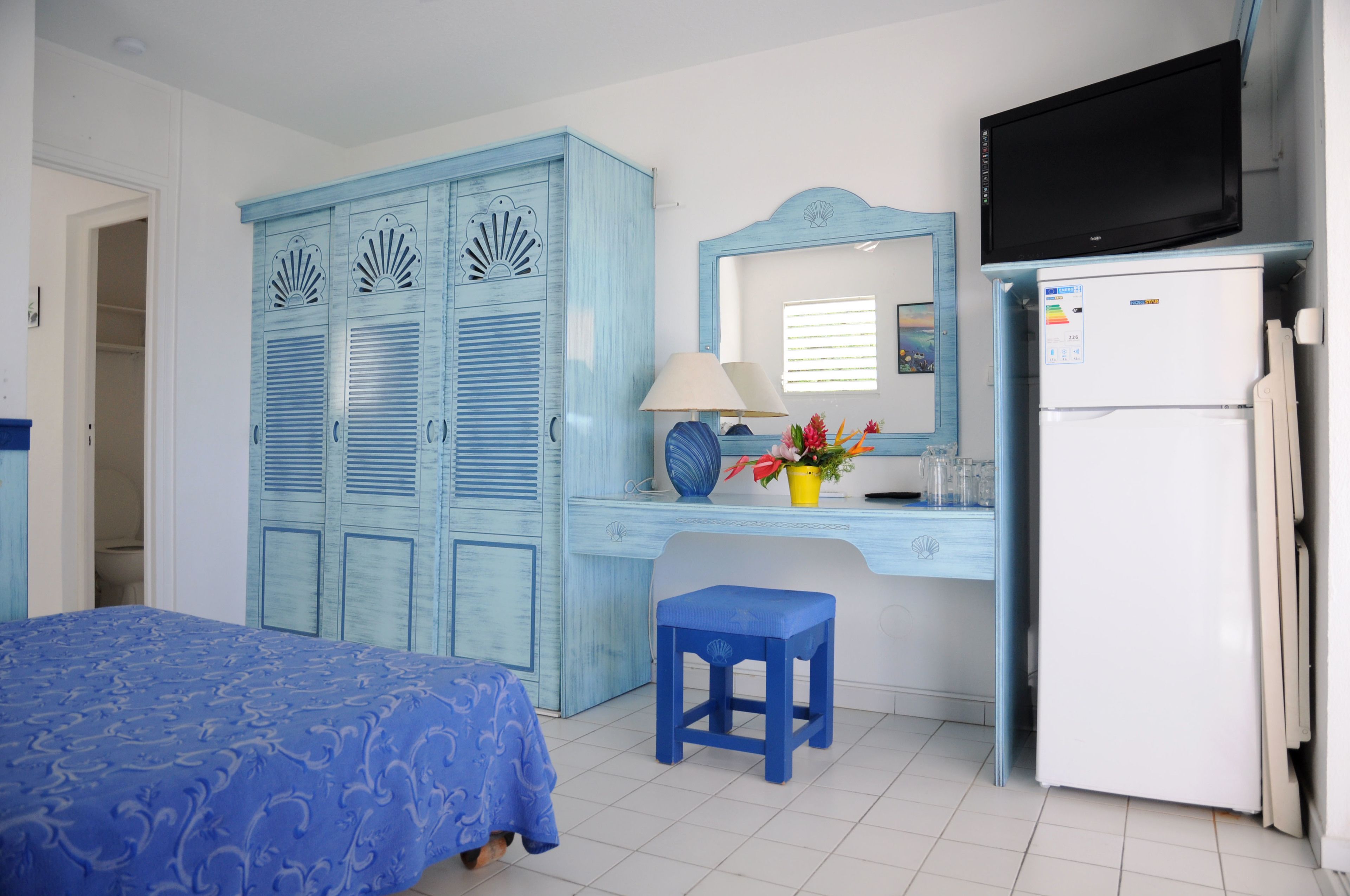 Quarto com cama, guarda-roupa azul, escrivaninha com espelho, televisão e frigorífico.