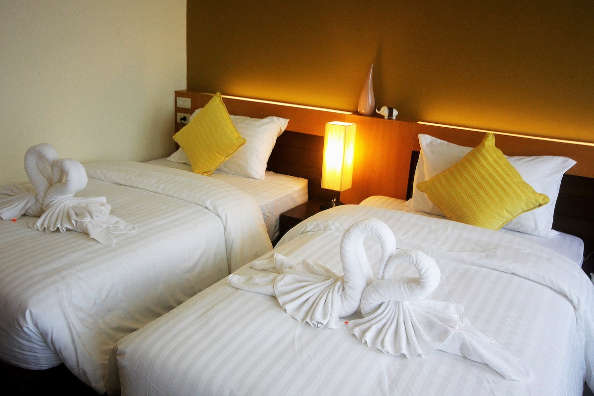 Duas camas de hotel com lençóis brancos e toalhas em formato de cisne, almofadas amarelas e um abajur.