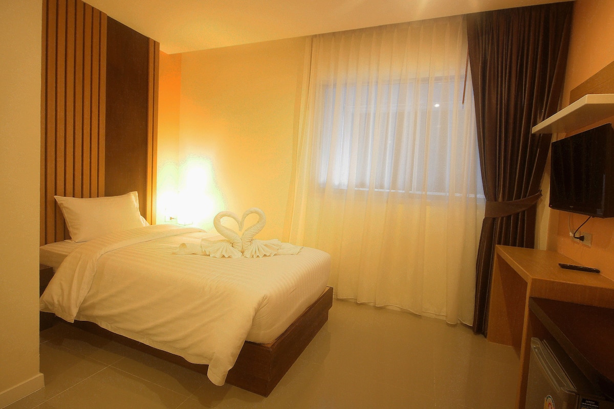 Cama bem arrumada com roupa de cama branca e arranjo de toalhas em formato de coração, cabeceira de madeira, mesa de cabeceira com controle remoto e janela com cortinas leves.