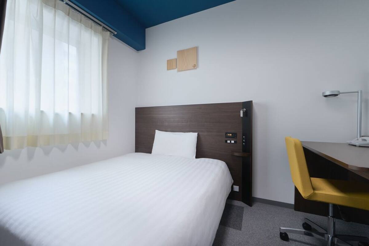 Quarto de hotel com uma cama, uma escrivaninha, uma cadeira, um abajur e uma janela com cortinas.