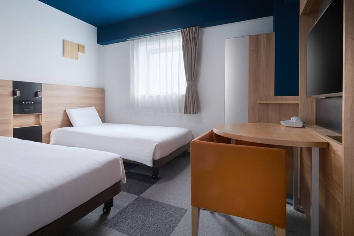 Quarto de hotel com duas camas, uma escrivaninha, uma cadeira, uma televisão e uma janela com cortinas.