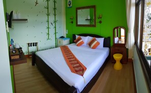 Cama bem arrumada com travesseiros e cobertor, mesa de cabeceira com abajur, escrivaninha com computador e janela com vista para o exterior.