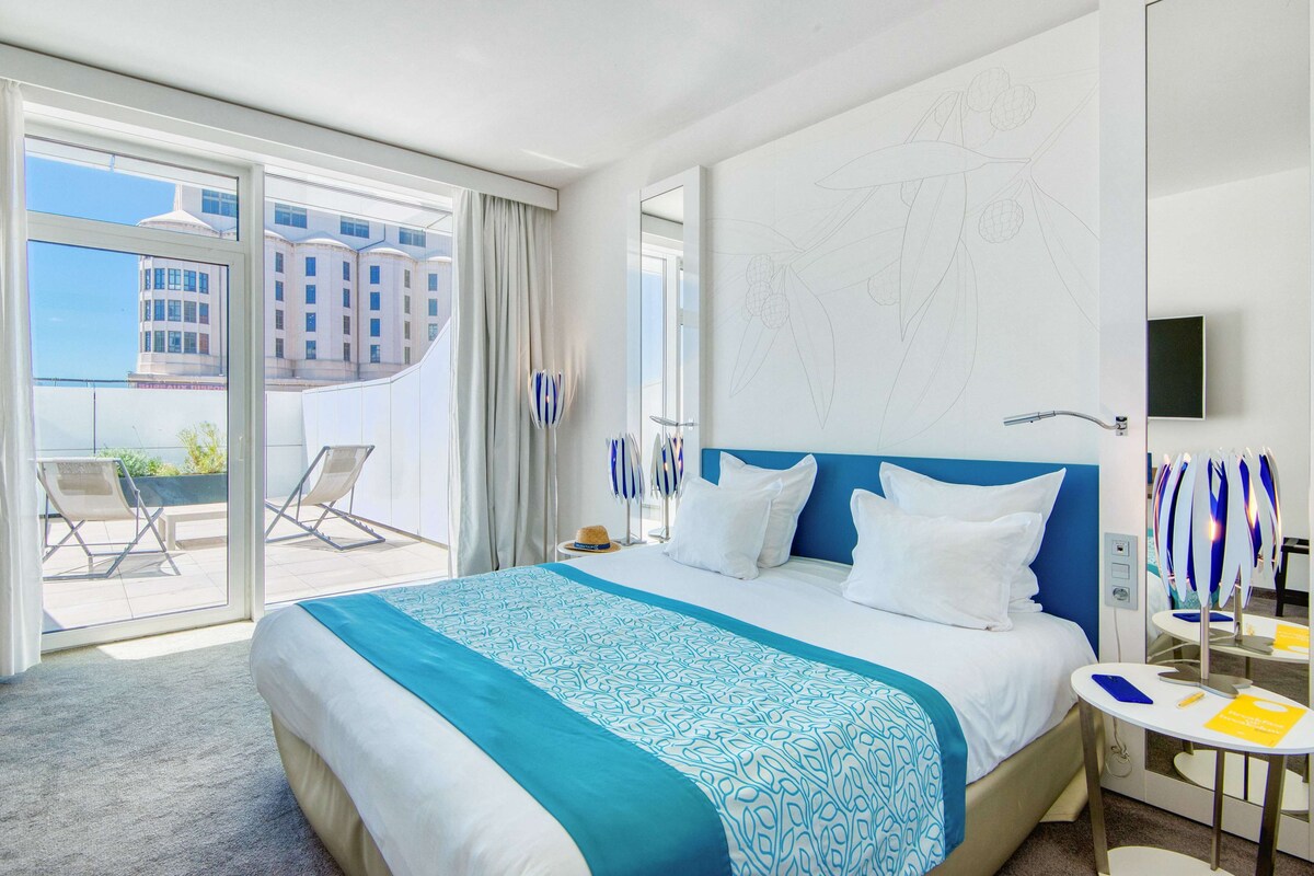 Quarto de hotel com uma cama grande, varanda com cadeiras e vista para um edifício.