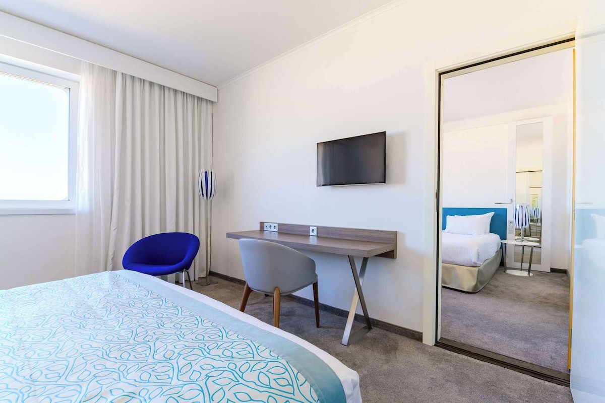 Quarto de hotel com uma cama, uma mesa, uma cadeira, uma televisão e um espelho.
