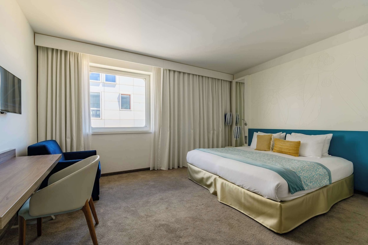 Quarto de hotel com cama, escrivaninha, cadeira e janela com cortinas.