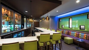 Uma área de bar com banquetas verdes, um balcão comprido e uma área de estar com parede estampada e colorida.