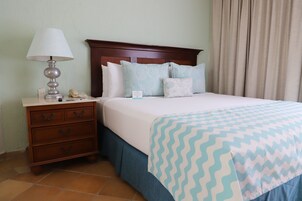 Uma cama bem arrumada com colcha estampada, uma mesa de cabeceira com abajur e uma mesinha de madeira.