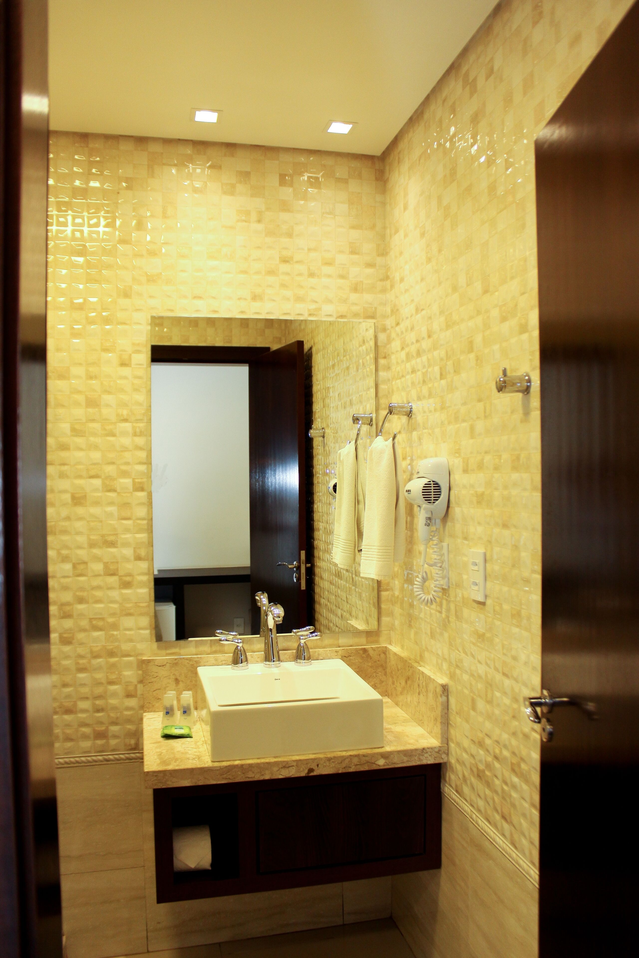 Um banheiro com parede revestida de azulejos bege, pia com espelho, porta-toalhas e secador de cabelo.
