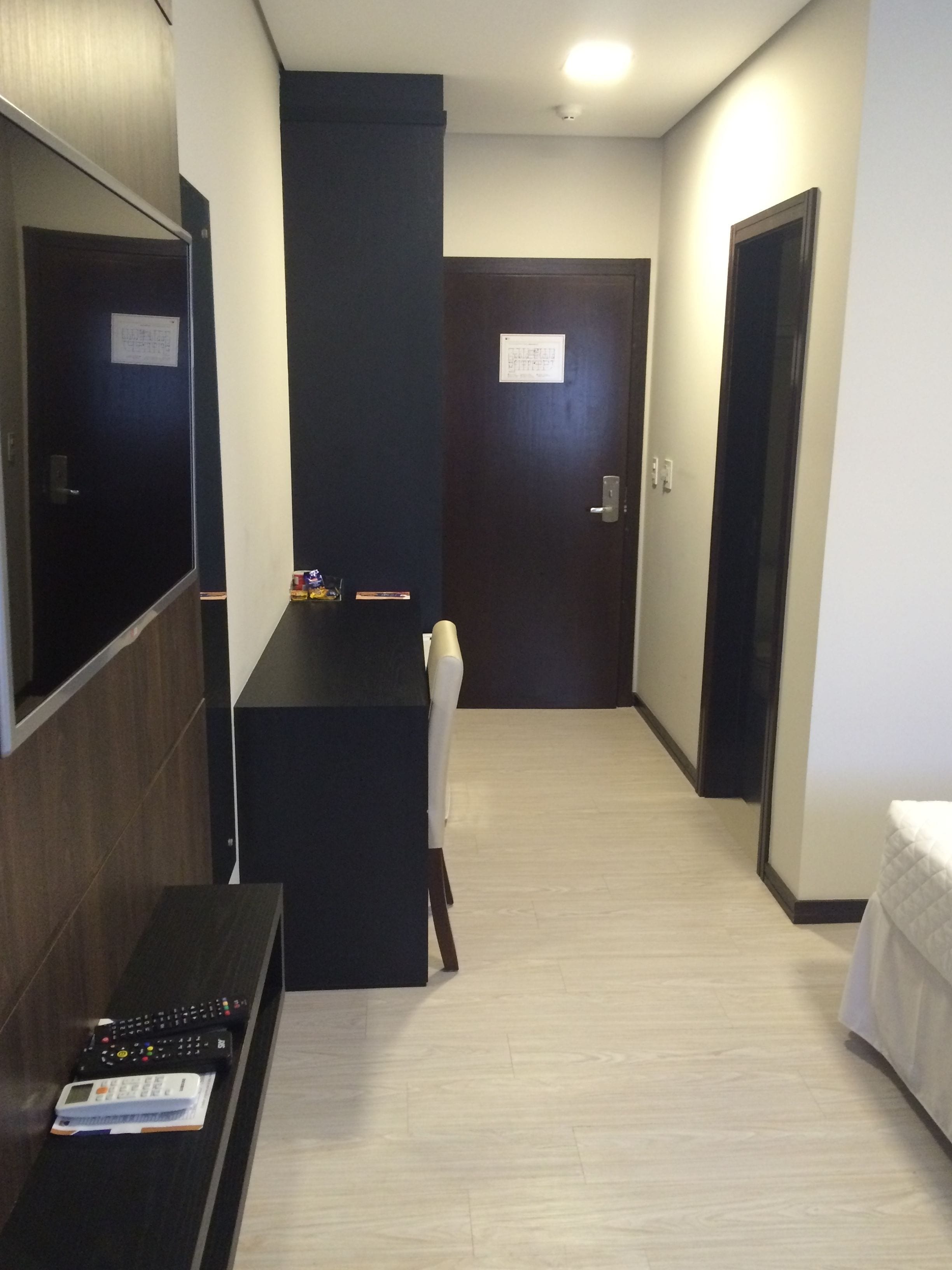 Quarto de hotel com uma cama, escrivaninha, cadeira, televisão e porta.