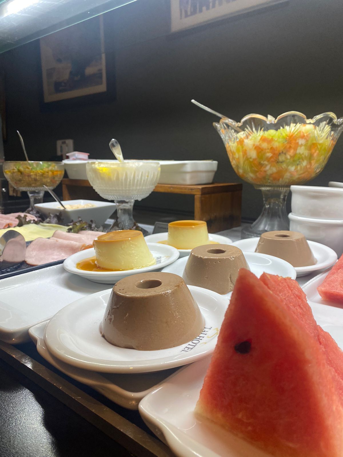 Buffet com diversos pratos, incluindo uma grande saladeira, vários doces e uma fatia de melancia.