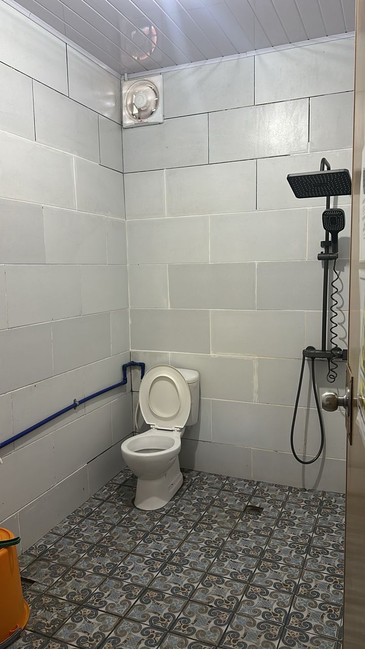 Casa de banho pequena com paredes revestidas a azulejos brancos, sanita, chuveiro e barra de apoio azul.