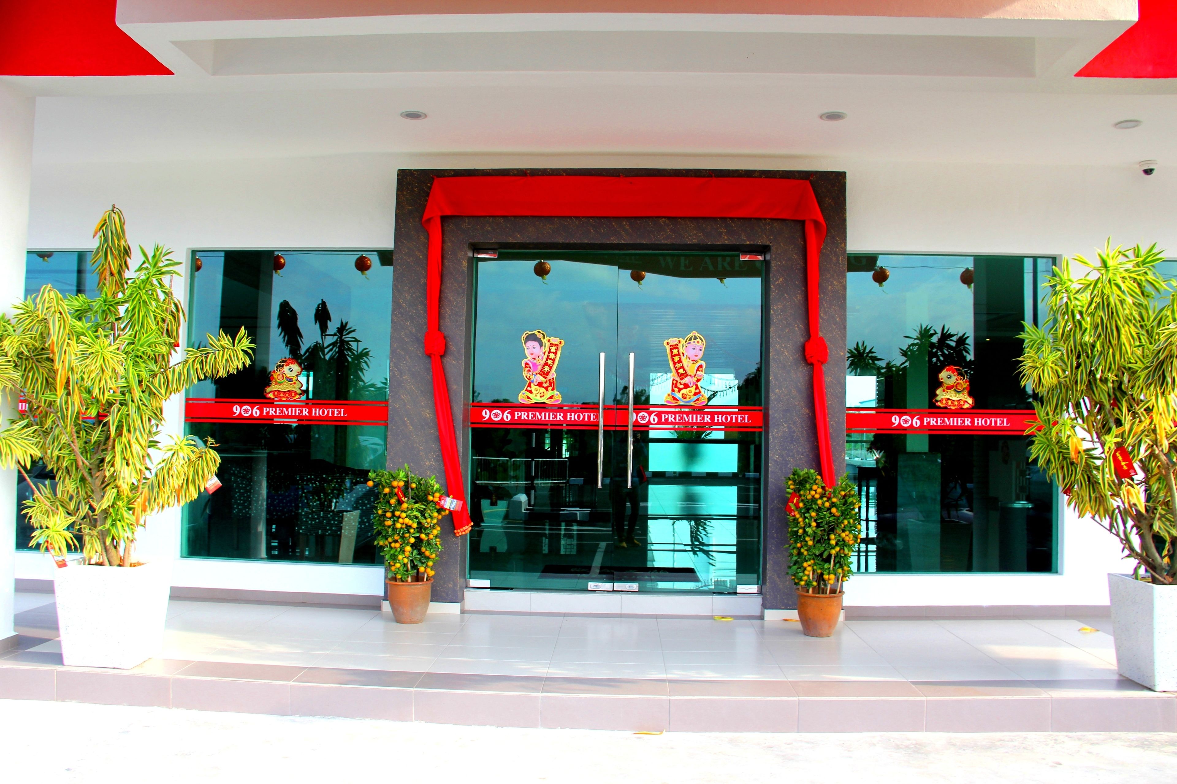 Exterior do Premier Hotel 906, com toldo vermelho, grandes janelas de vidro e vasos de plantas.