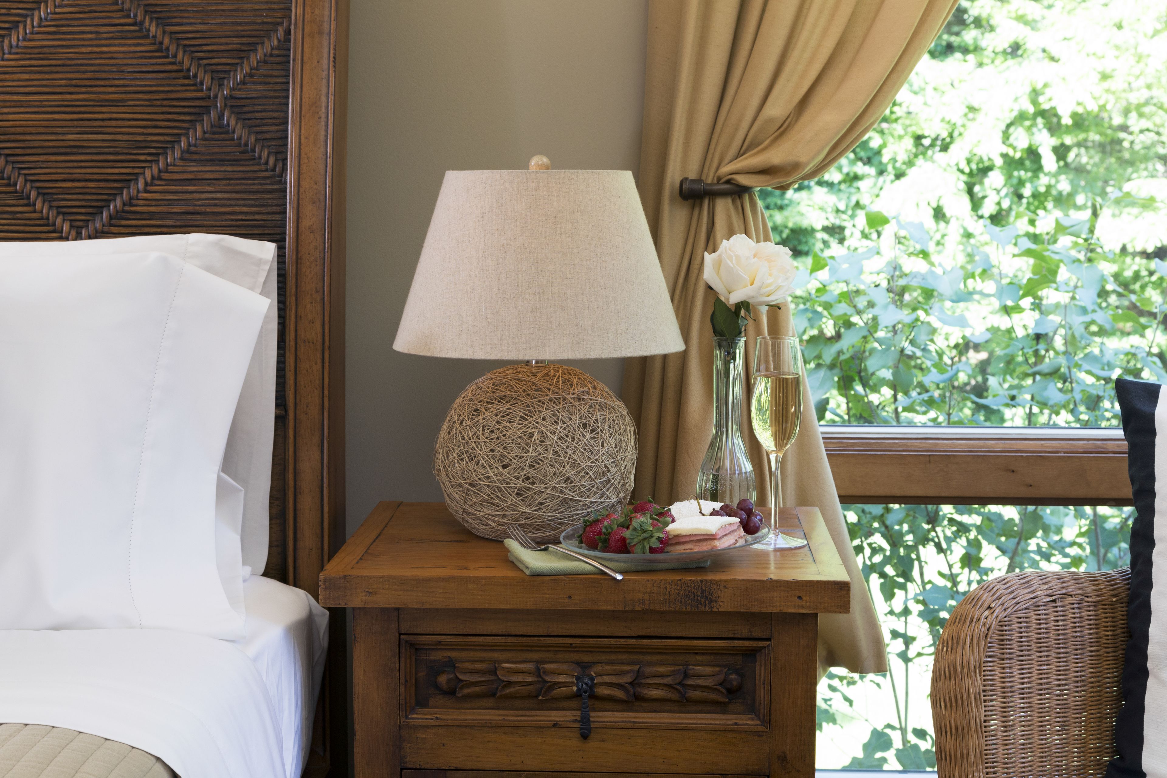 Cama bem arrumada com lençóis brancos, mesa de cabeceira de madeira com abajur, bola de vime, vaso de vidro com flor branca e prato com frutas e queijo.