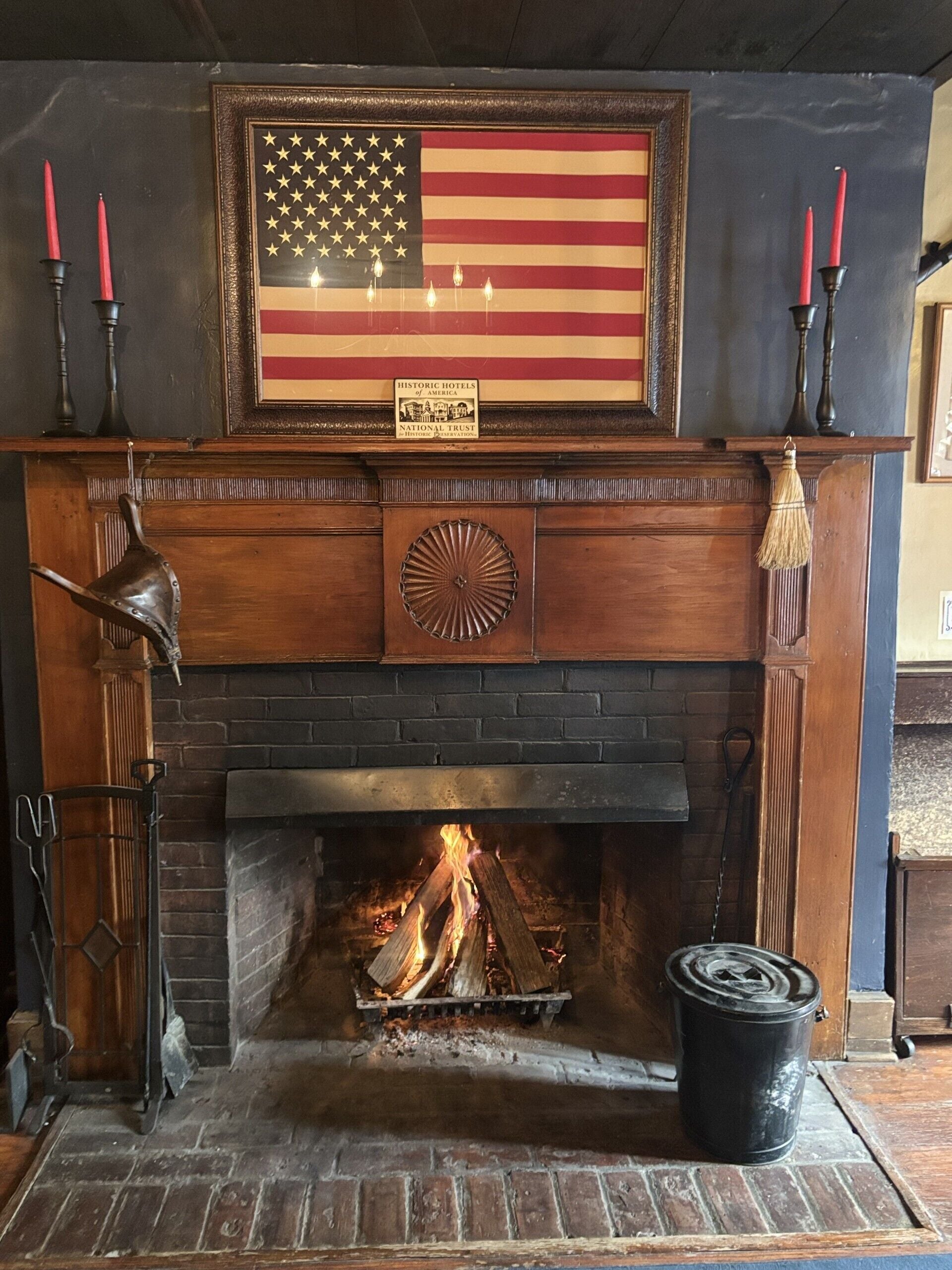 Uma lareira com uma bandeira americana emoldurada, uma placa indicando ser um hotel histórico e uma panela preta no braseiro.