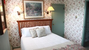 Quarto com cama de madeira, papel de parede floral, duas luminárias na parede e um quadro emoldurado sobre a cama.