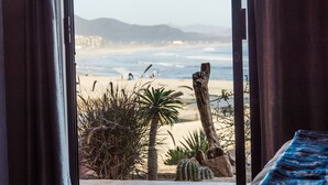 Vista de um quarto com janela para uma praia, mostrando um poste de madeira, plantas típicas do deserto e uma cadeia de montanhas ao longe.