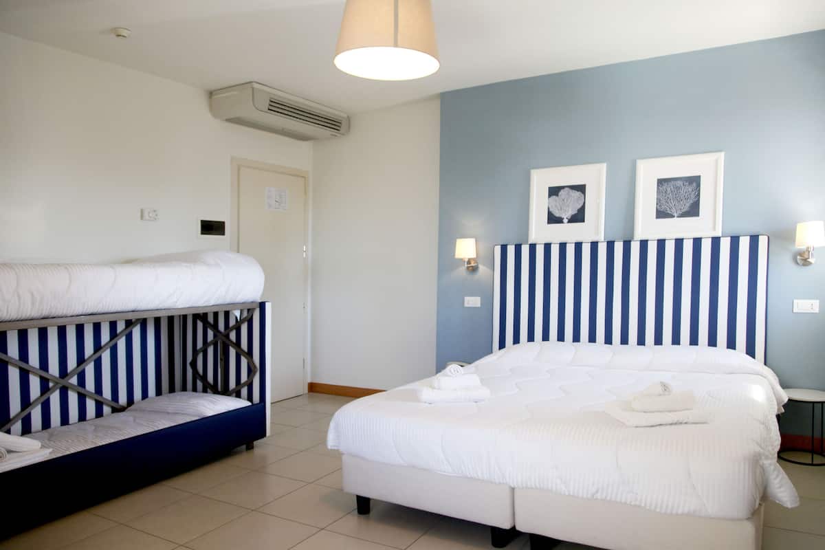 Quarto de hotel com cama beliche, uma cama de solteiro e duas luminárias fixadas na parede.