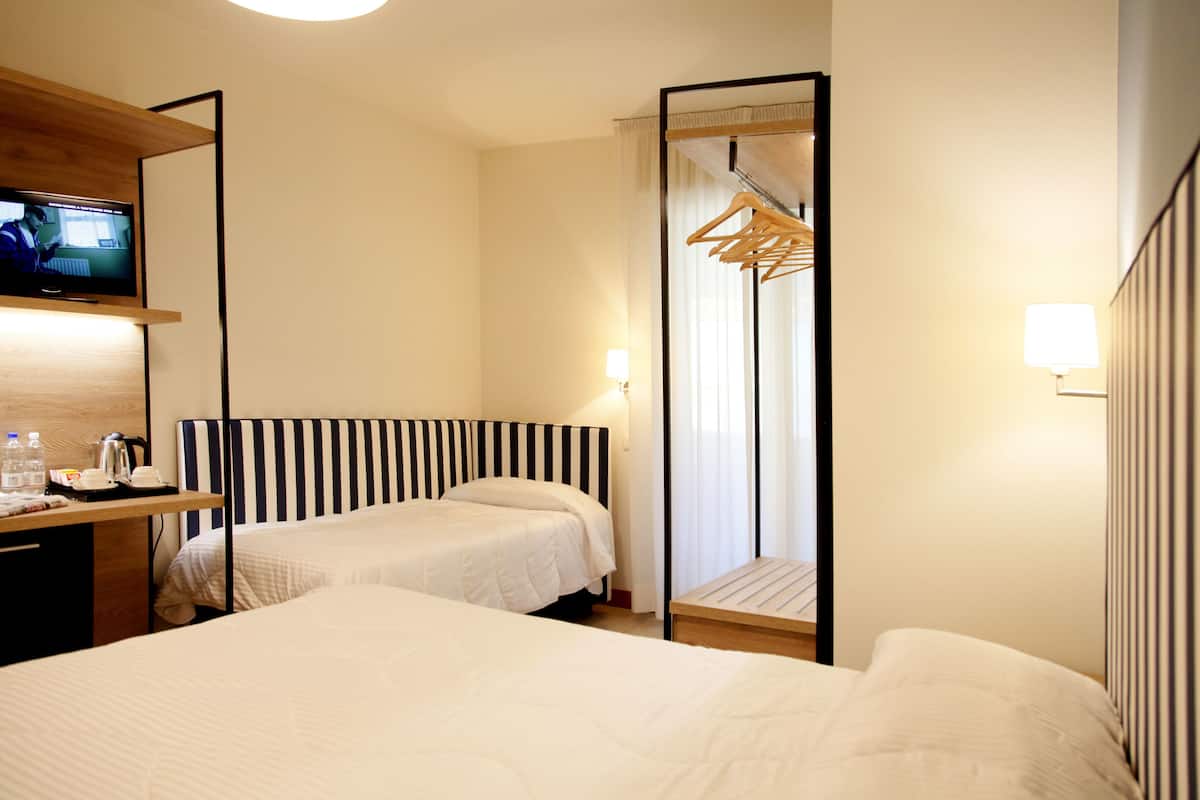 Quarto de hotel com duas camas, uma TV em uma prateleira, uma escrivaninha com chaleira e um cabideiro.