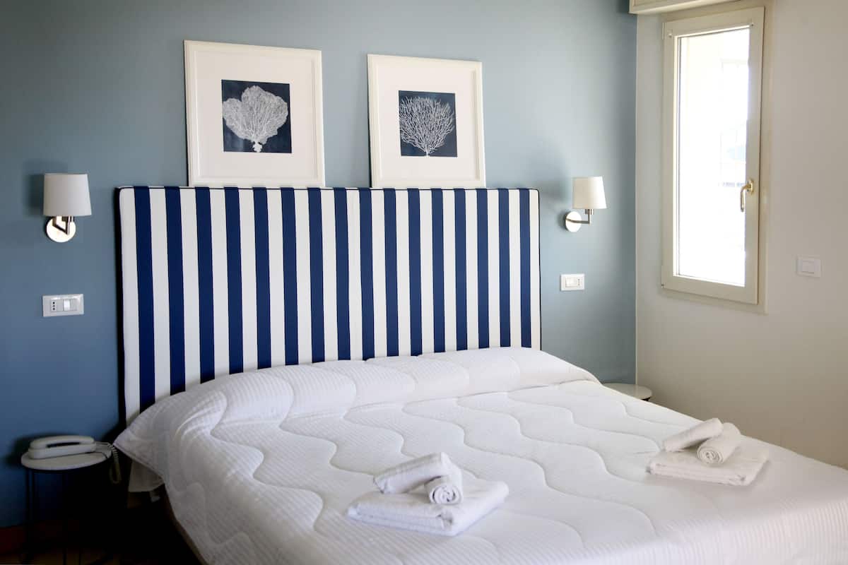 Cama bem arrumada com roupa de cama branca e toalhas dobradas, cabeceira listrada, duas obras de arte emolduradas e luminárias de cabeceira.
