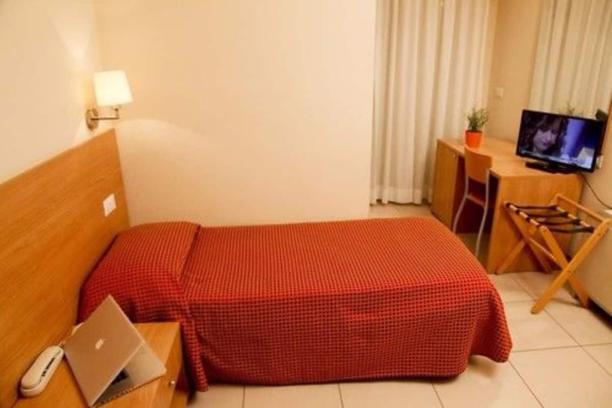 Quarto de hotel com uma cama de solteiro, uma mesa com computador, uma televisão e uma pequena mesa com uma planta.
