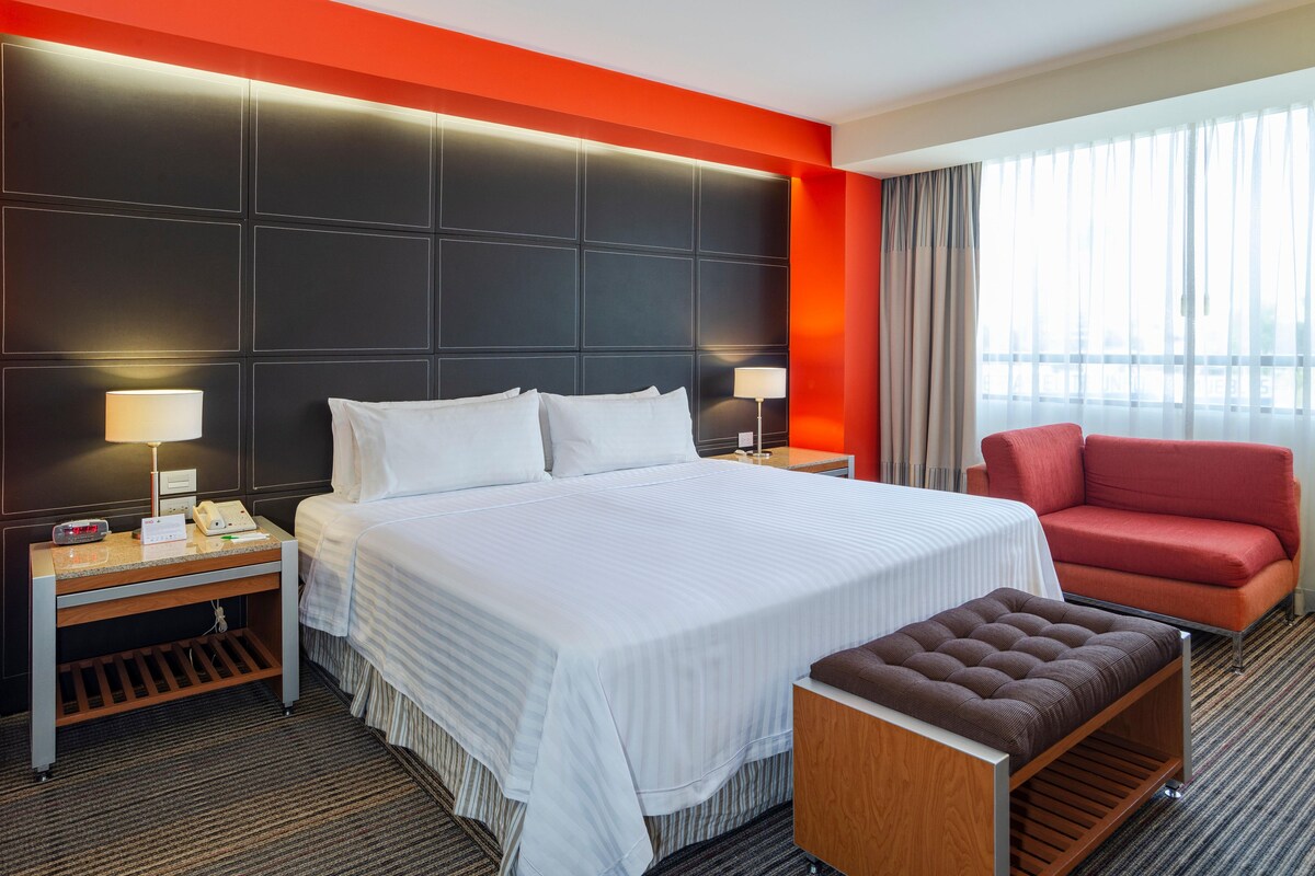 Quarto de hotel com uma cama grande, mesas de cabeceira, uma poltrona vermelha e um banco de madeira.