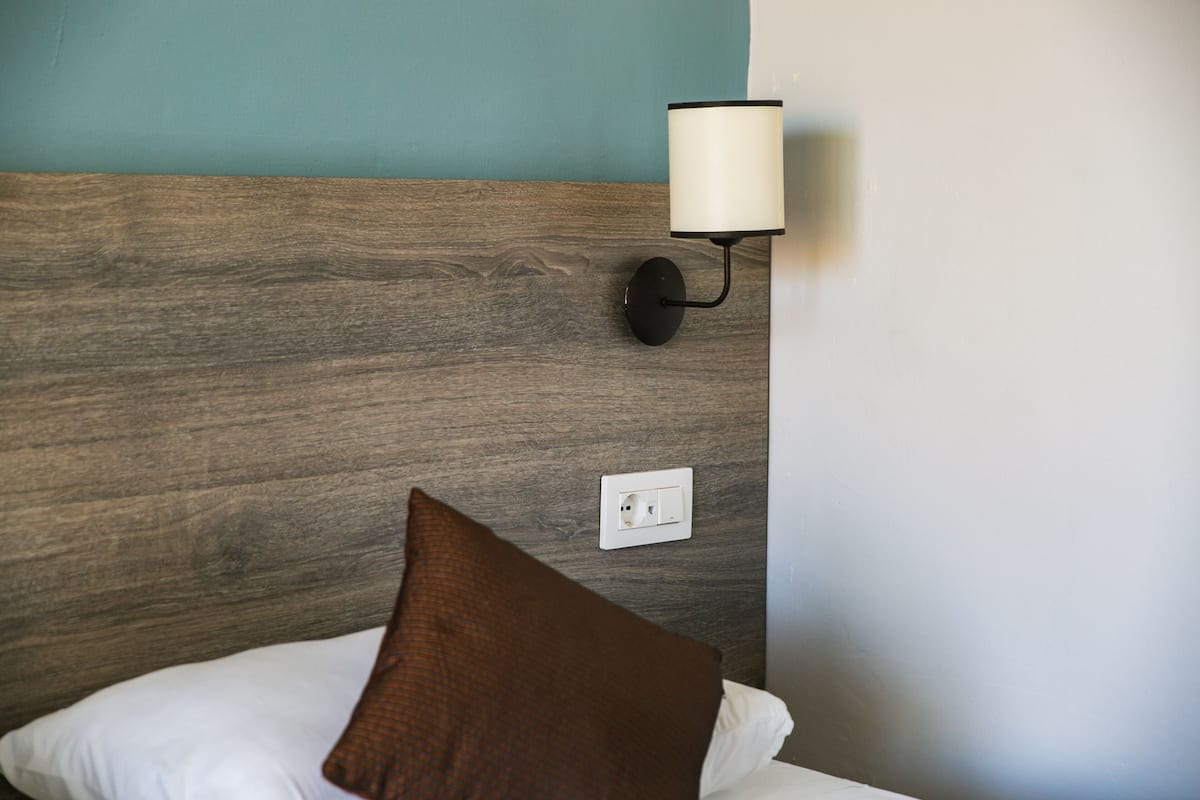 Quarto de hotel com cabeceira de madeira, luminária fixada na parede, tomada e travesseiro castanho na cama.