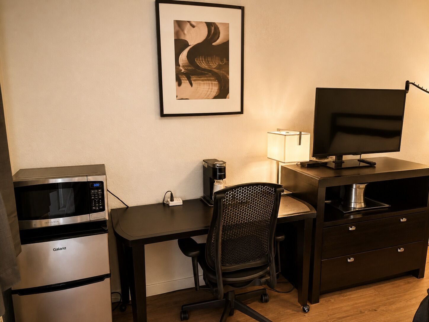 Quarto de hotel com cozinha compacta, escrivaninha com cadeira, televisão de tela plana e uma obra de arte emoldurada na parede.