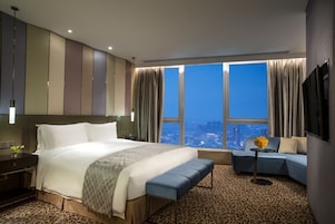 Quarto de hotel moderno com uma cama grande, um sofá, uma mesinha e vista para a cidade através de grandes janelas.