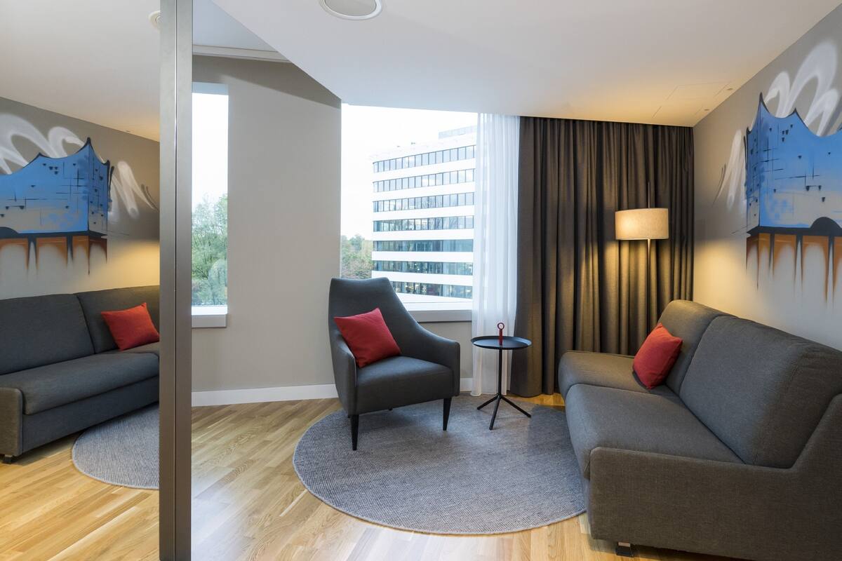 Quarto de hotel moderno com sofá cinzento, um travesseiro vermelho, uma mesinha redonda e uma luminária. Há uma janela grande com vista para um edifício e árvores.