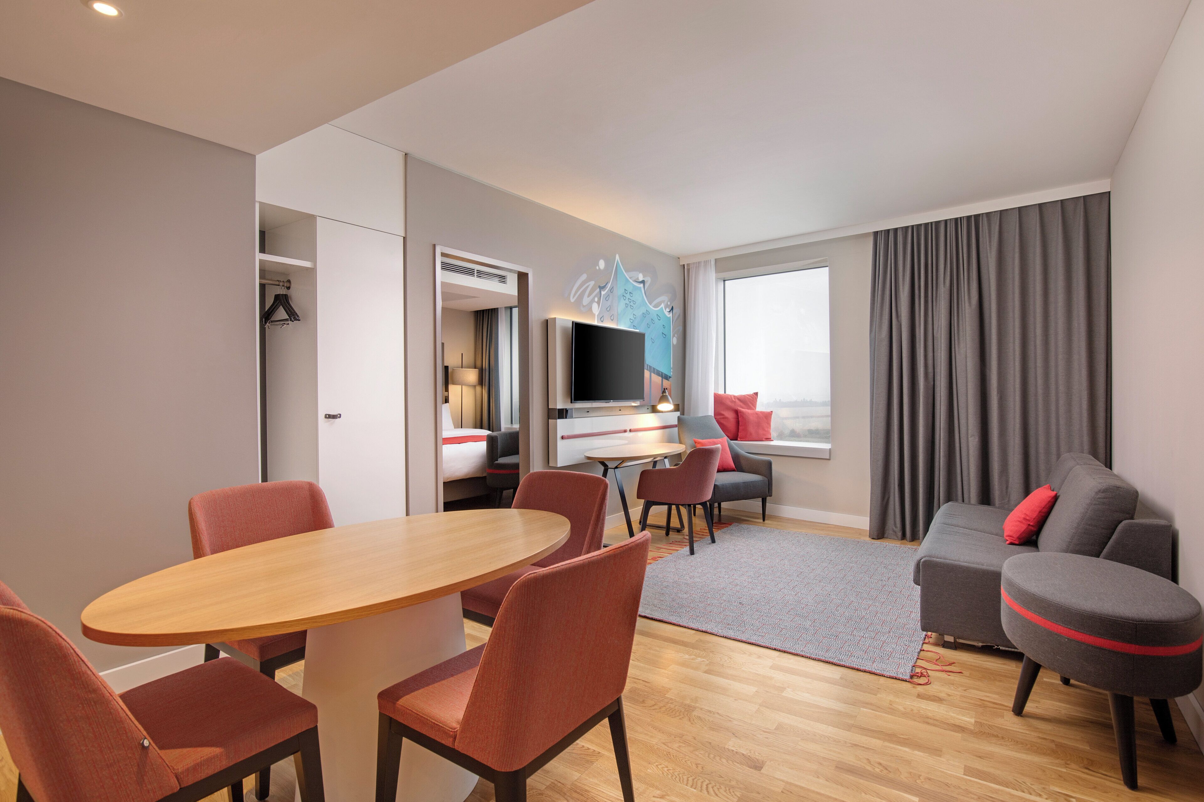 Quarto de hotel moderno com área de jantar, sofá, televisão e uma janela grande com cortinas.