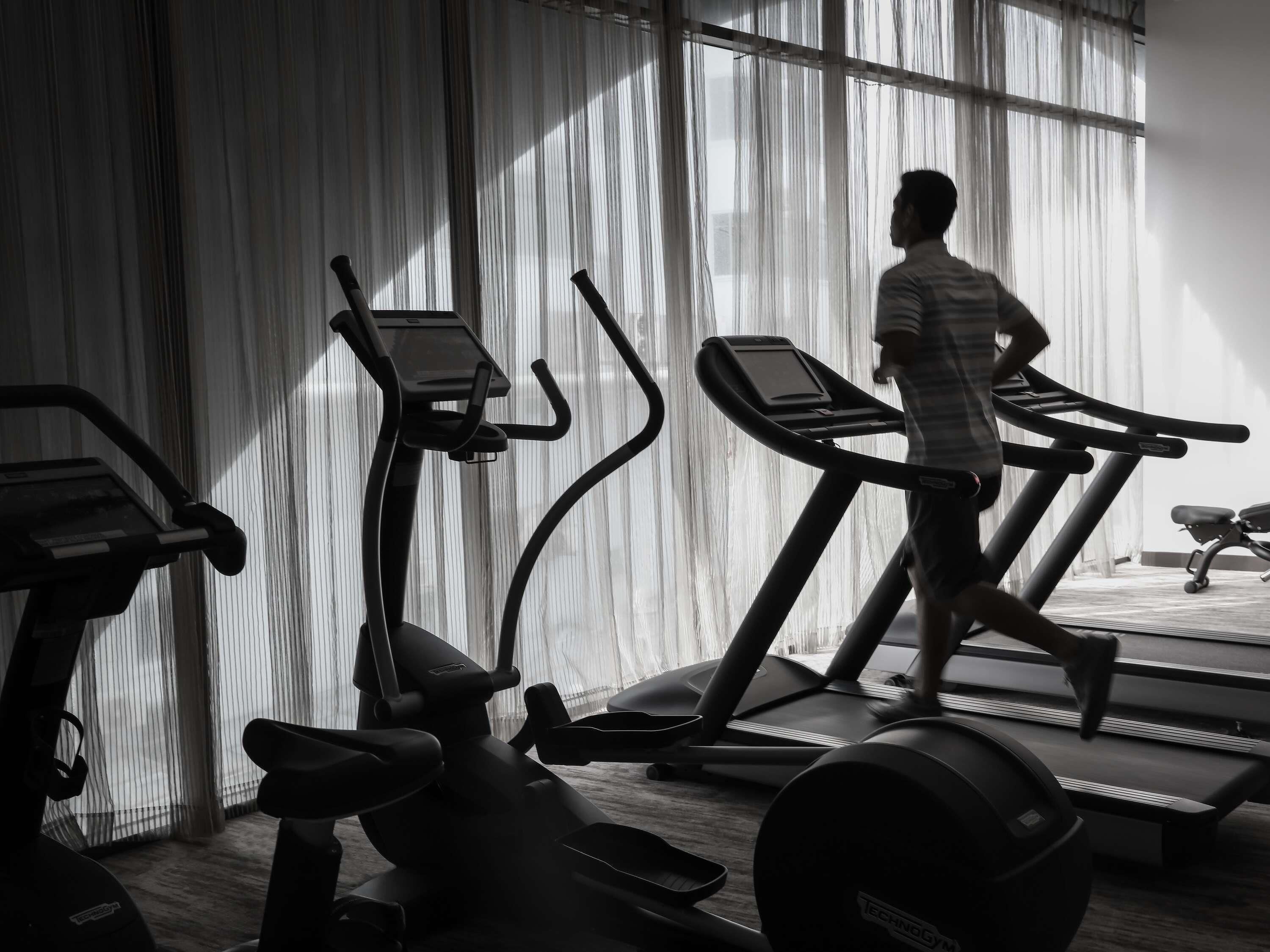 Uma academia com esteiras e equipamentos de exercício, grandes janelas com cortinas leves, e uma pessoa caminhando em uma esteira.