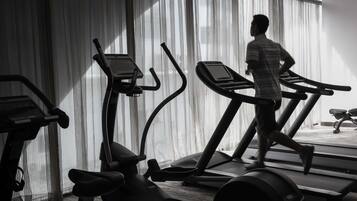 Uma academia com esteiras e equipamentos de exercício, grandes janelas com cortinas leves, e uma pessoa caminhando em uma esteira.