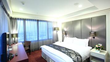 Quarto de hotel com uma cama grande, uma escrivaninha com computador, uma cadeira, mesa de cabeceira com abajur e vista para a cidade pelas janelas.